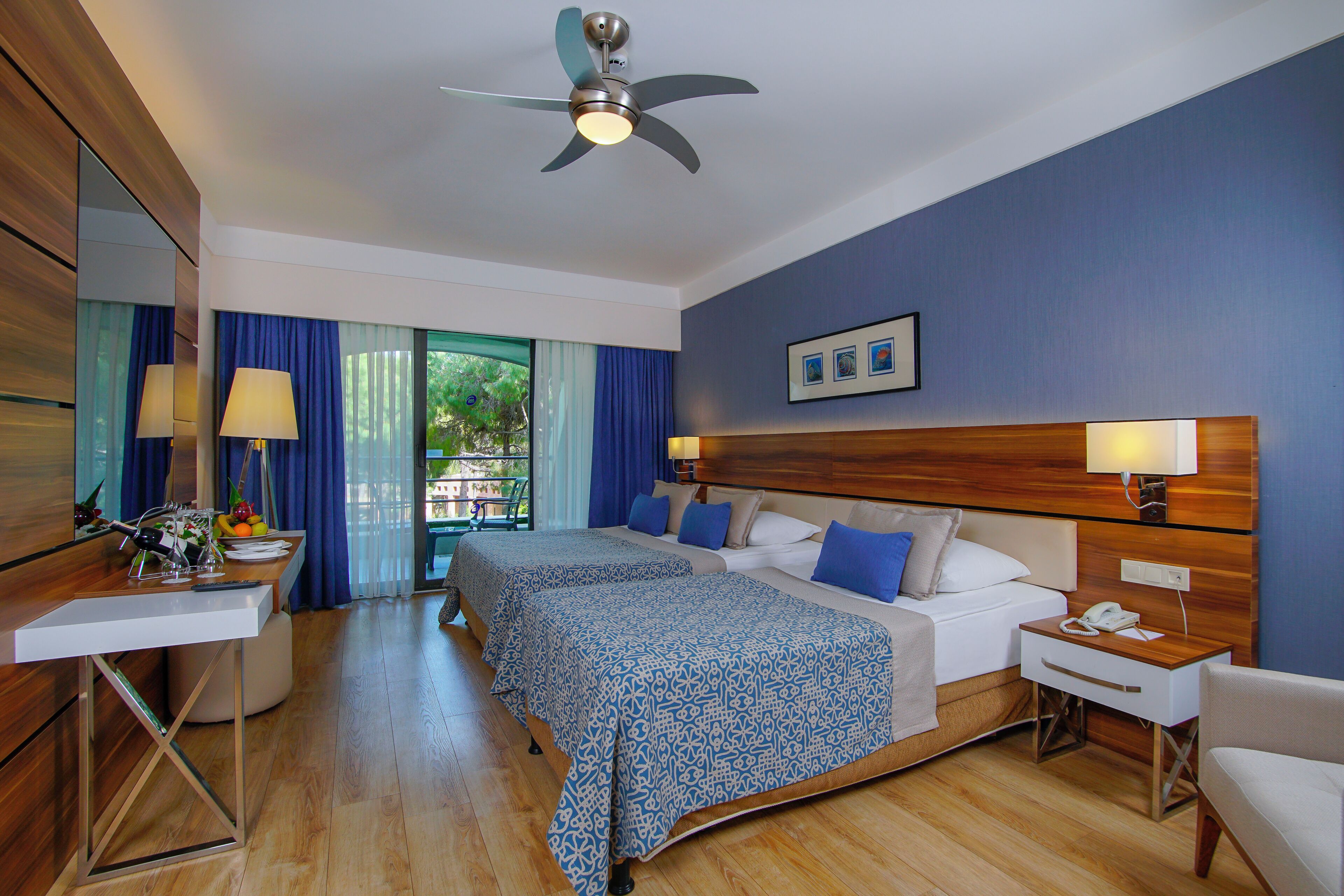 Quarto de hotel com duas camas, uma escrivaninha com luminária, uma cadeira, mesa de cabeceira com telefone e ventilador de teto.