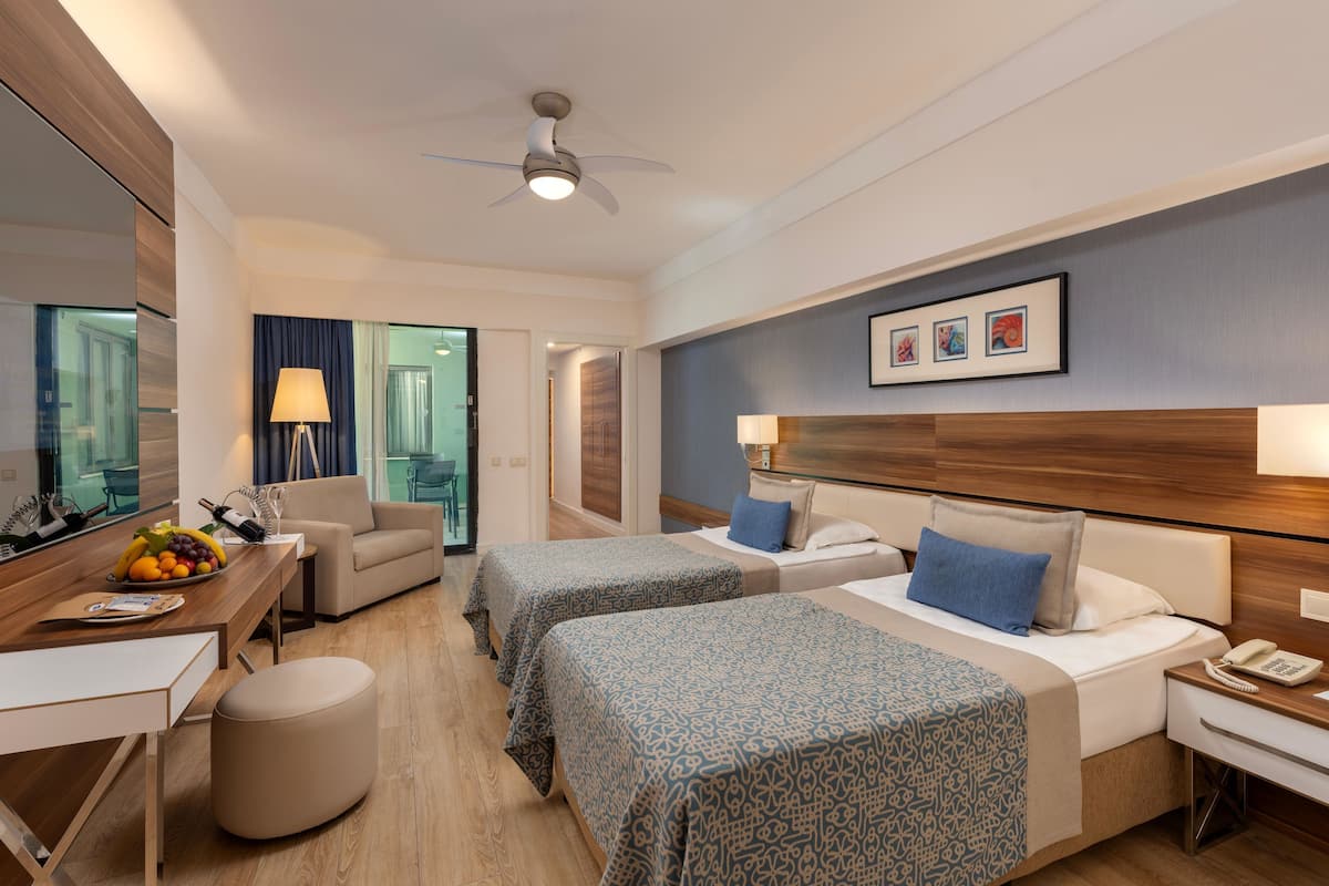 Quarto de hotel com duas camas, uma escrivaninha, uma cadeira, uma mesinha com fruta e um ventilador de teto.