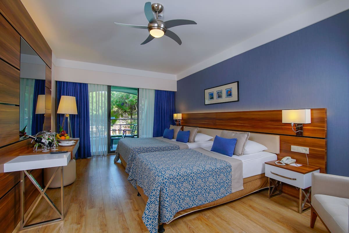 Quarto de hotel com duas camas, uma escrivaninha, uma cadeira, um ventilador de teto e uma janela com cortinas.