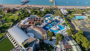 Vista aérea de um resort com uma grande piscina central, várias piscinas menores e uma área de praia com guarda-sóis.