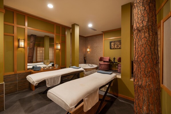 Sala de spa com duas macas de massagem, uma banheira e um espelho grande.