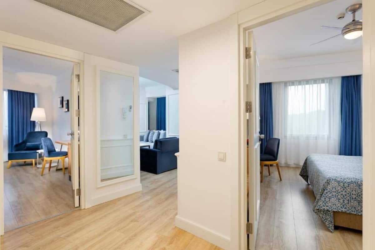 Quarto de hotel com uma cama, uma escrivaninha e uma cadeira. Há uma sala de estar com um sofá e um abajur.