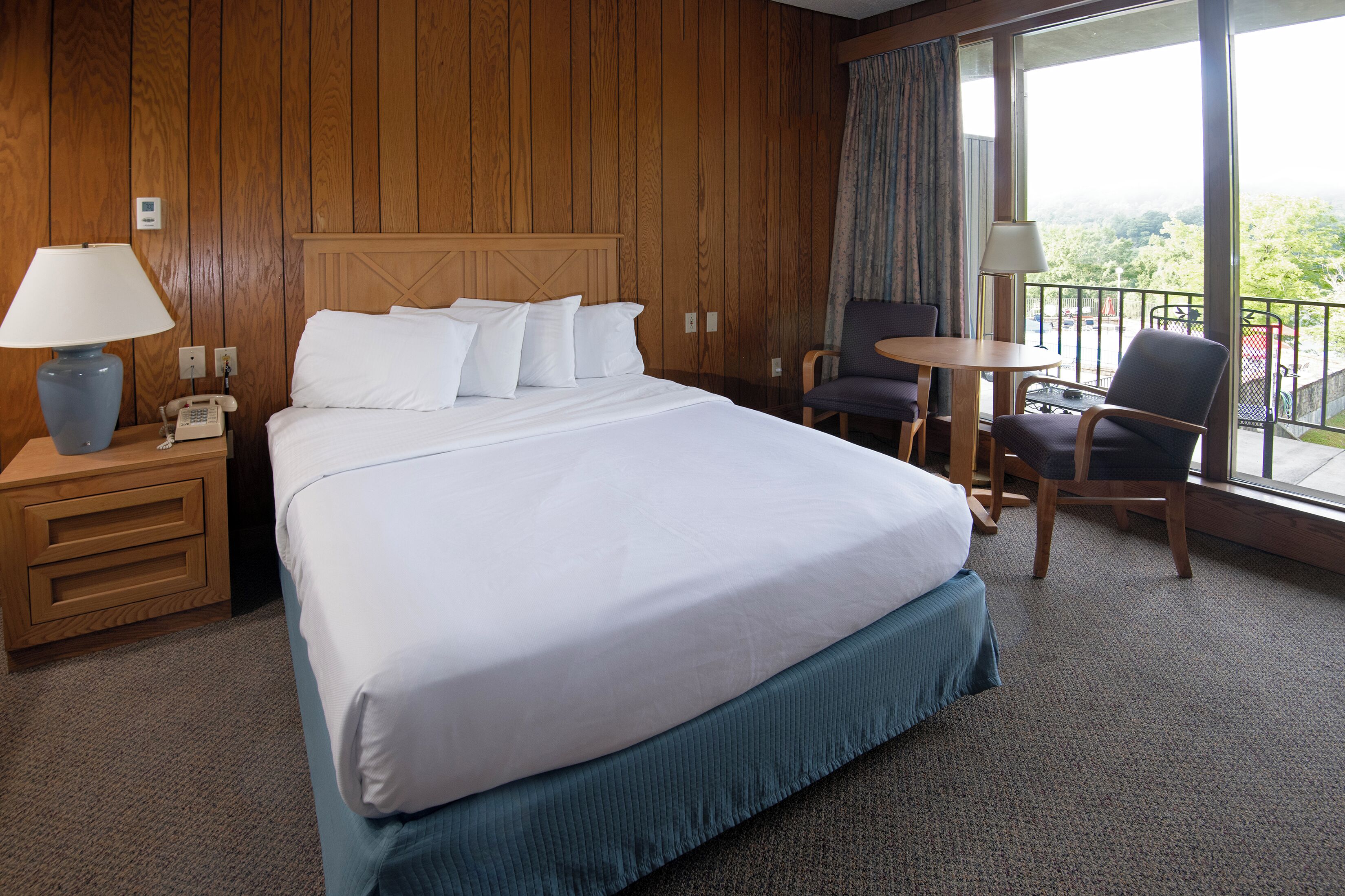 Quarto de hotel com uma cama grande, cabeceira de madeira, mesa de cabeceira com abajur, pequena mesa com cadeiras e vista para o exterior por grandes janelas.