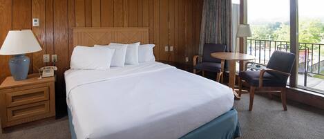 Quarto de hotel com uma cama grande, cabeceira de madeira, mesa de cabeceira com abajur, pequena mesa com cadeiras e vista para o exterior por grandes janelas.