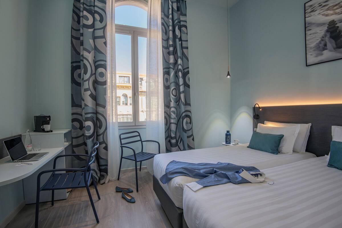 Quarto de hotel com cama, escrivaninha com laptop, cadeira e vista para edifícios.