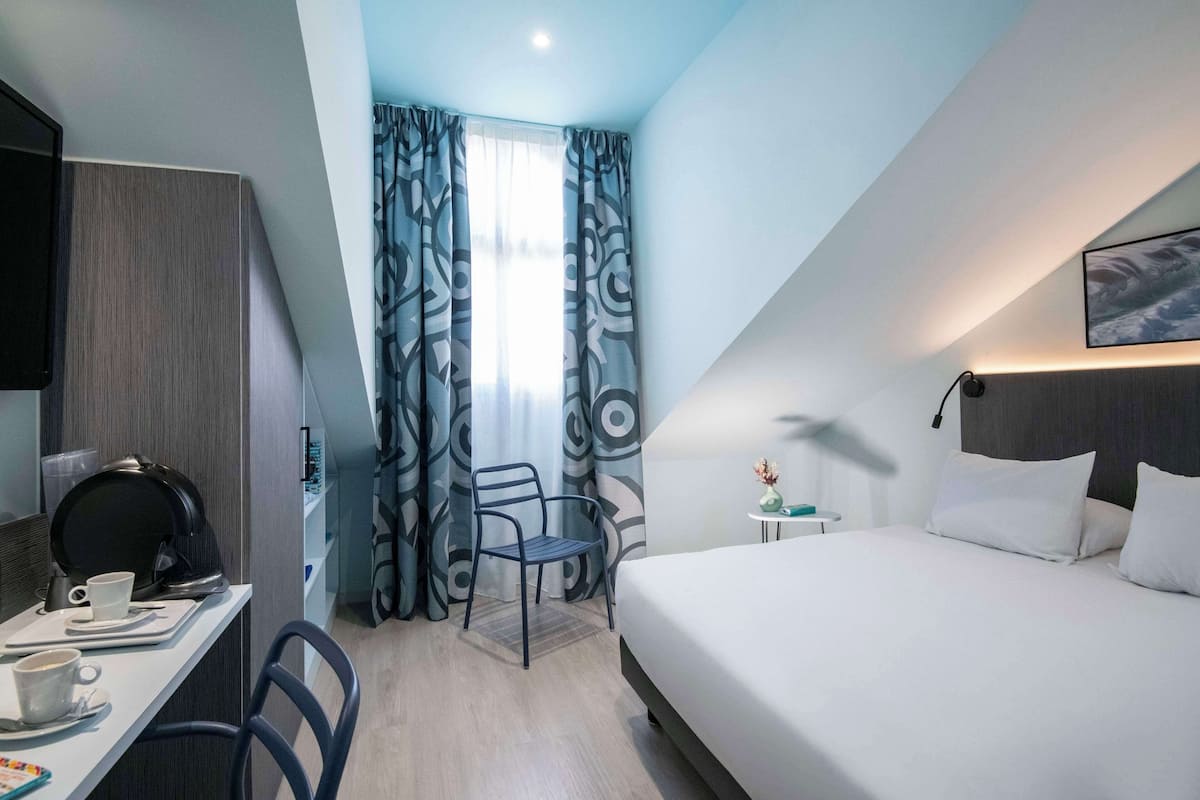 Quarto de hotel moderno com cama, escrivaninha com cadeira, mesa pequena com abajur e janela com cortinas.