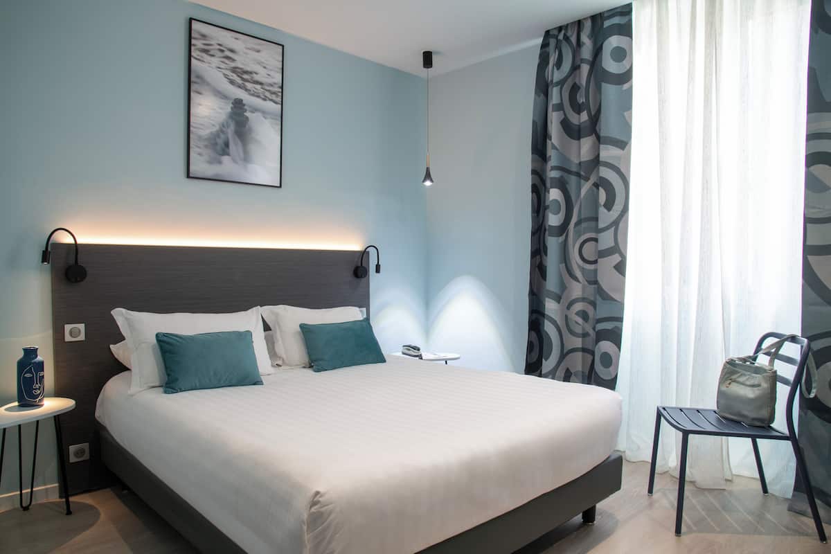 Quarto de hotel moderno com uma cama grande, uma mesinha de cabeceira com luminária, uma cadeira com uma bolsa e uma janela com cortinas.