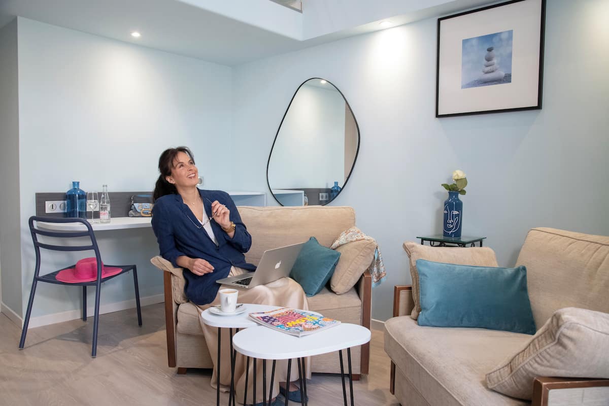 Quarto de hotel moderno com sofá, uma mesinha com um laptop, uma cadeira e uma xícara de café.