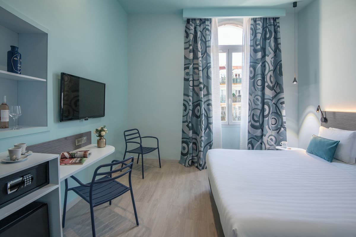 Quarto de hotel com cama, escrivaninha com cadeira, televisão e janela com cortinas estampadas.
