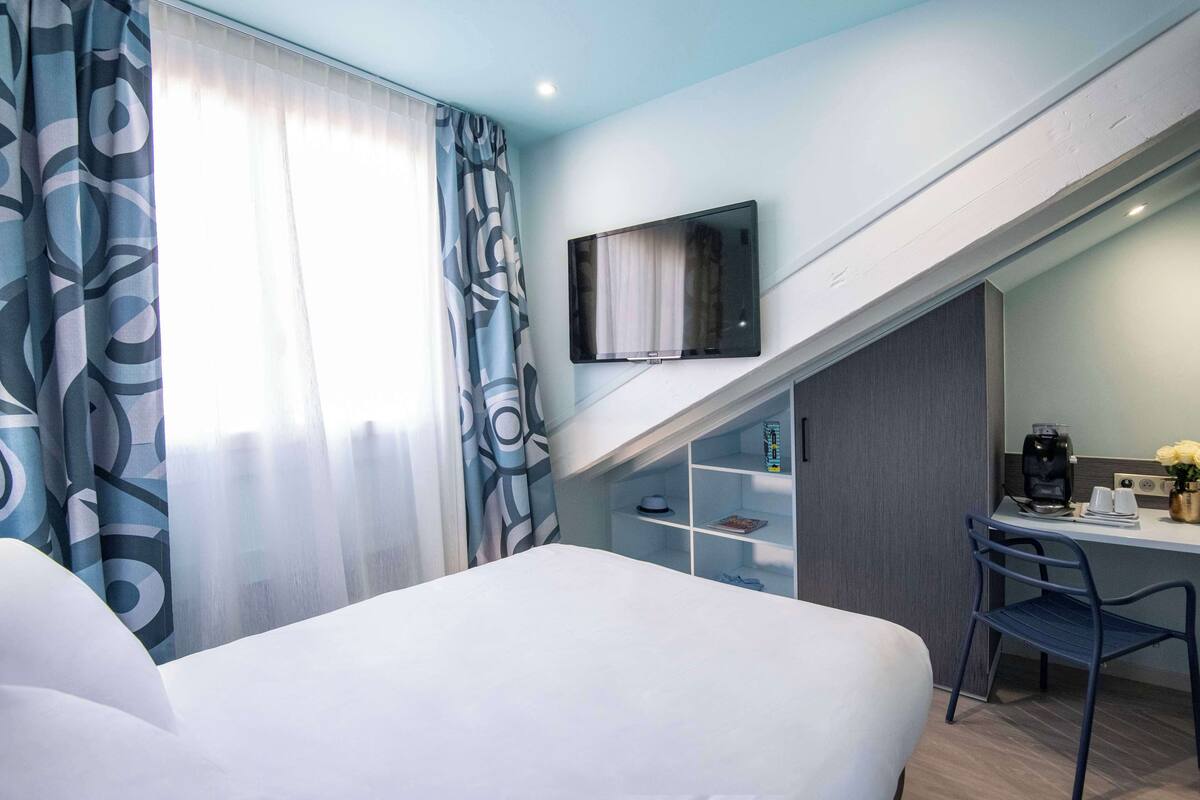 Quarto de hotel com cama, secretária com cadeira, televisão na parede e estante de livros.