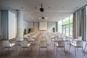 Uma sala de conferências com fileiras de cadeiras, uma tela de projetor e amplas janelas.