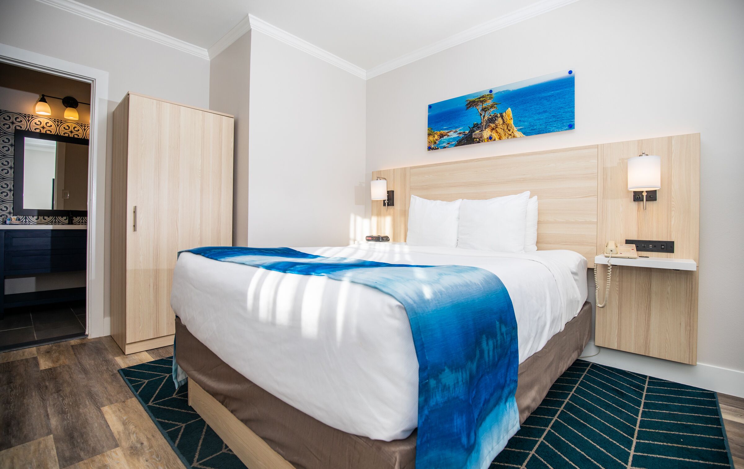 Quarto de hotel com uma cama grande, cabeceira de madeira, pintura representando uma paisagem costeira, mesa de cabeceira com abajur e um guarda-roupa.