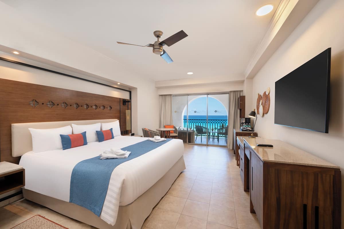 Quarto de hotel moderno com uma cama grande, ventilador de teto, televisão, varanda com vista para o mar e uma pequena área de estar com mesa e cadeiras.