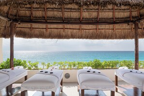 Um spa à beira-mar com telhado de palha, três mesas de massagem e vista para o oceano.