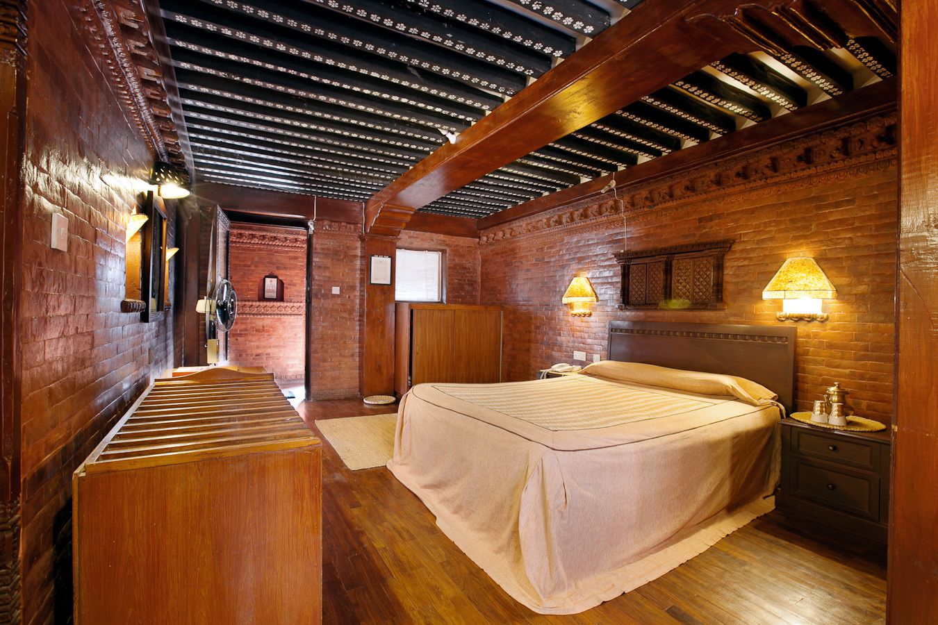 Quarto com piso de madeira, cama com cabeceira, mesa de cabeceira, abajur e parede de tijolos.