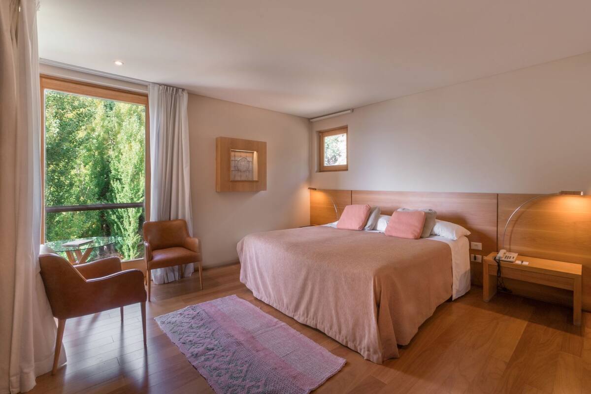 Quarto com cama grande, uma cadeira, mesa de cabeceira e janela com vista para árvores.