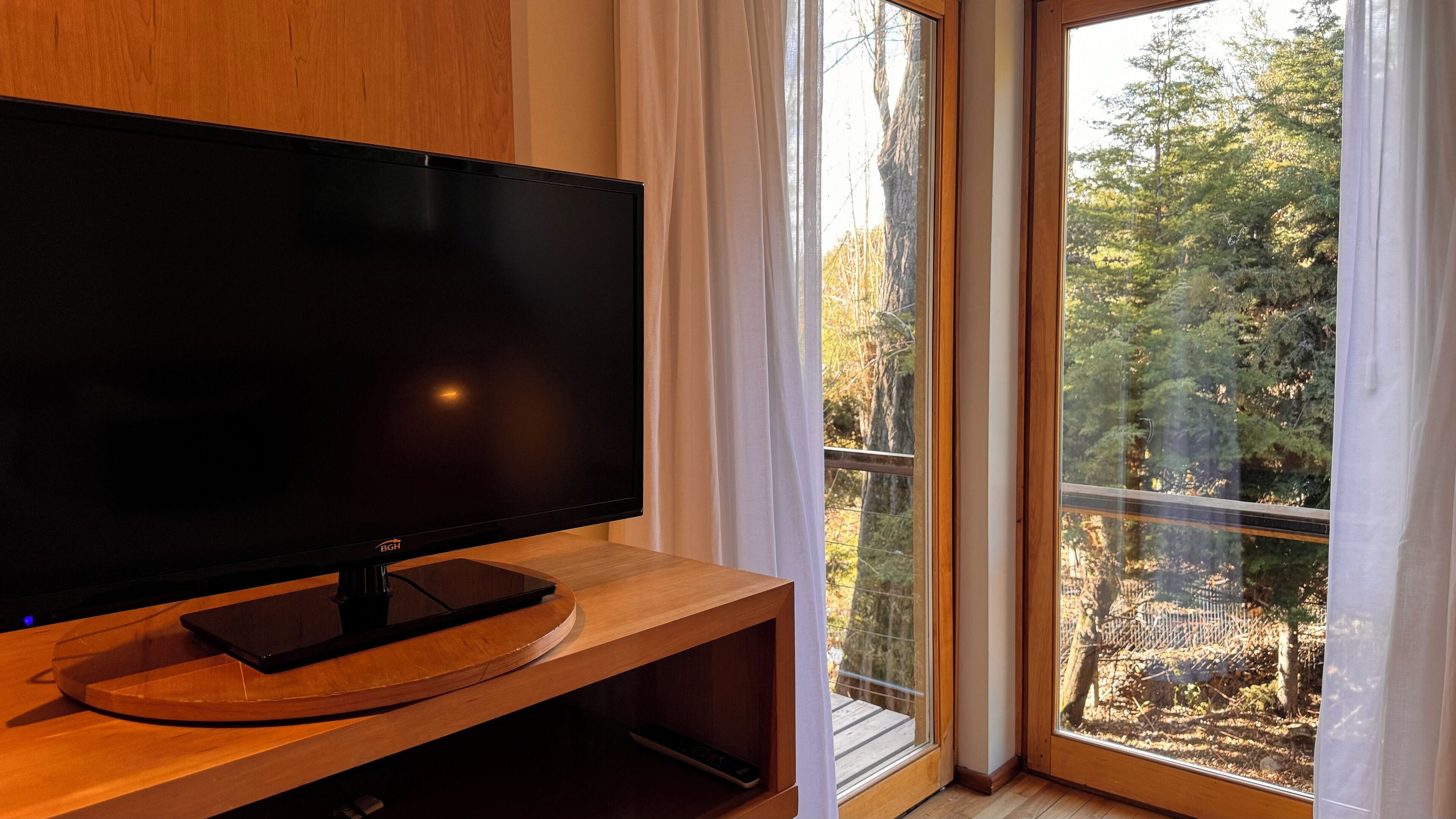 Televisão de tela plana em um suporte de madeira em um quarto com uma janela grande.