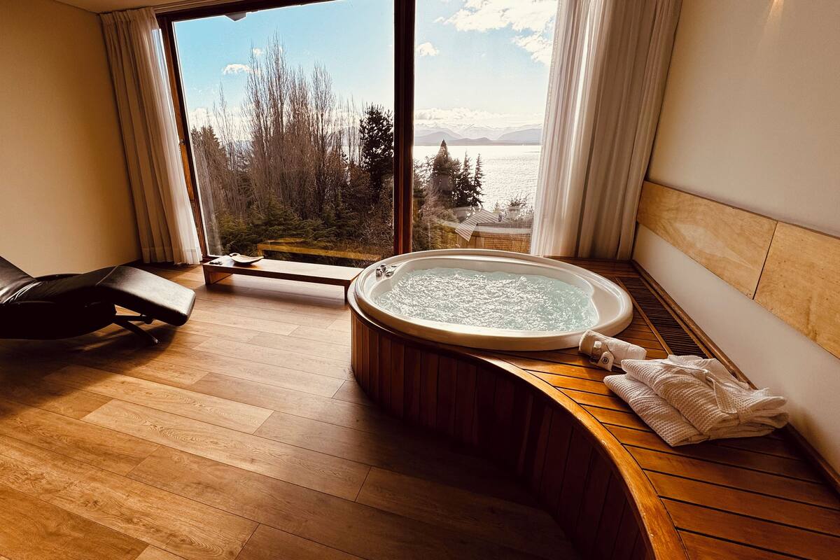 Um espaço de spa com piso de madeira, uma poltrona de descanso preta, uma banheira de hidromassagem e uma grande janela com vista para árvores e montanhas.