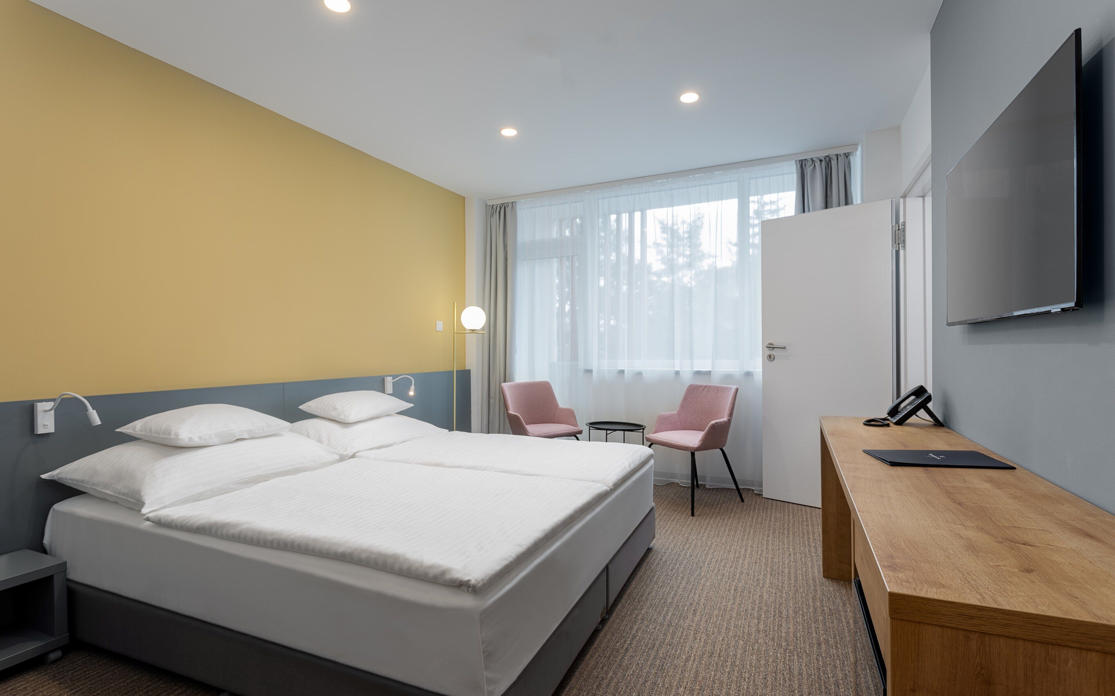 Quarto de hotel com uma cama grande, duas cadeiras, uma escrivaninha, televisão de tela plana e janela com cortinas.