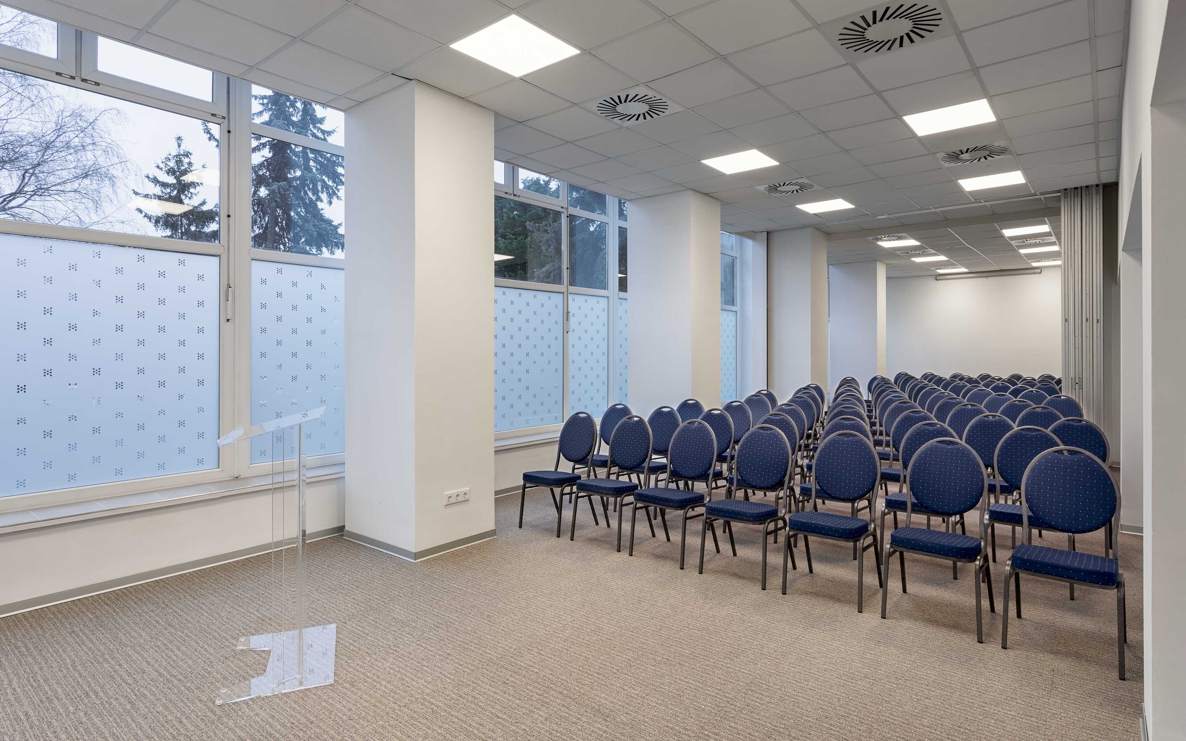 Sala de conferências com fileiras de cadeiras azuis, grandes janelas com vidro fosco e teto com iluminação embutida.