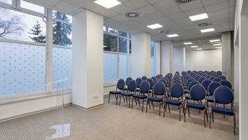 Sala de conferências com fileiras de cadeiras azuis, grandes janelas com vidro fosco e teto com iluminação embutida.