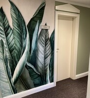 Um corredor com um mural na parede mostrando grandes folhas verdes e uma porta com a inscrição 'EAST'.