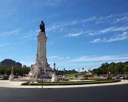 Um grande monumento com uma estátua no topo, rodeado por um espaço ajardinado com bancos e uma pequena fonte.