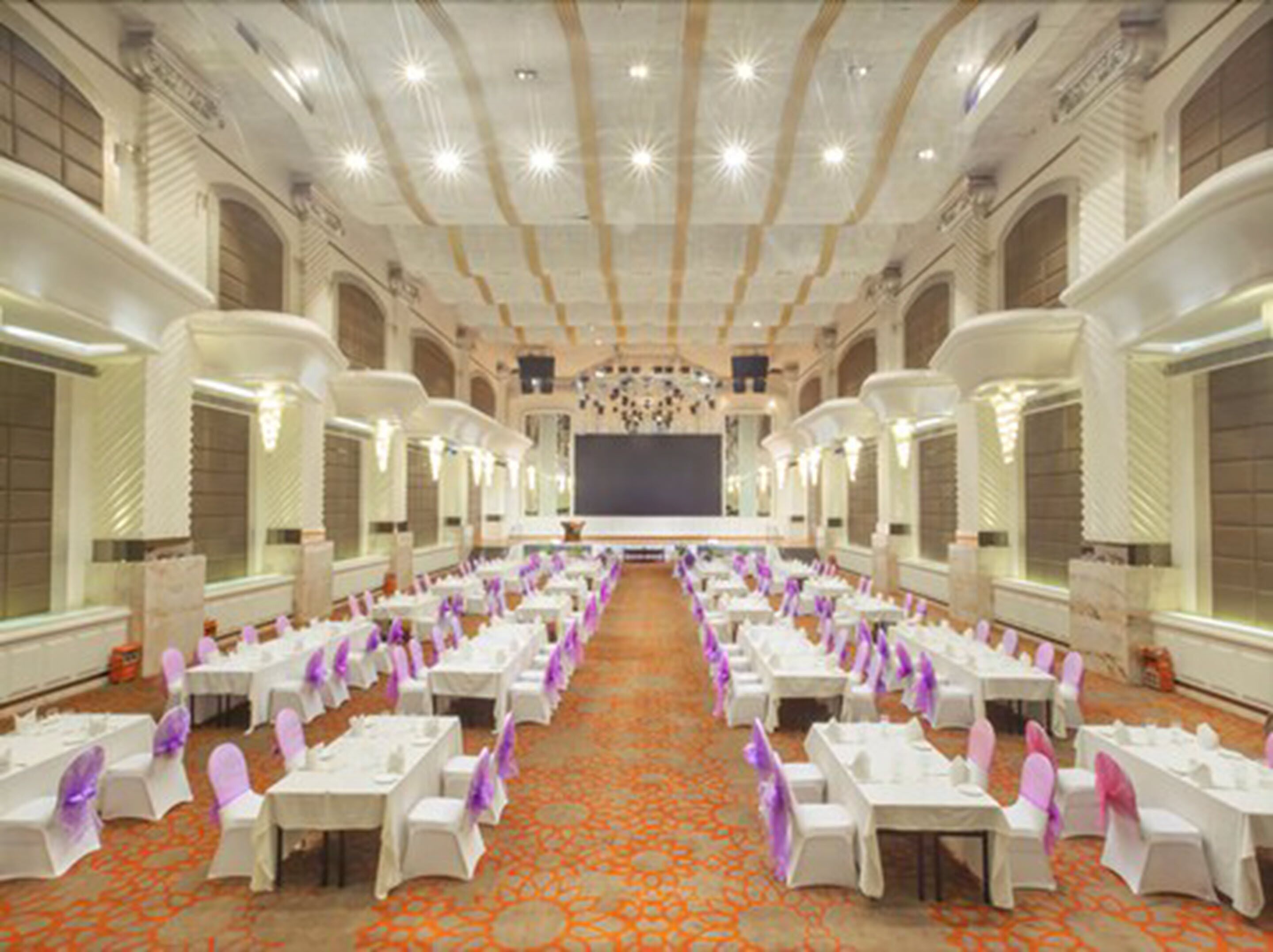 Uma sala de banquetes com fileiras de mesas e cadeiras, um palco na frente e detalhes arquitetônicos elaborados.
