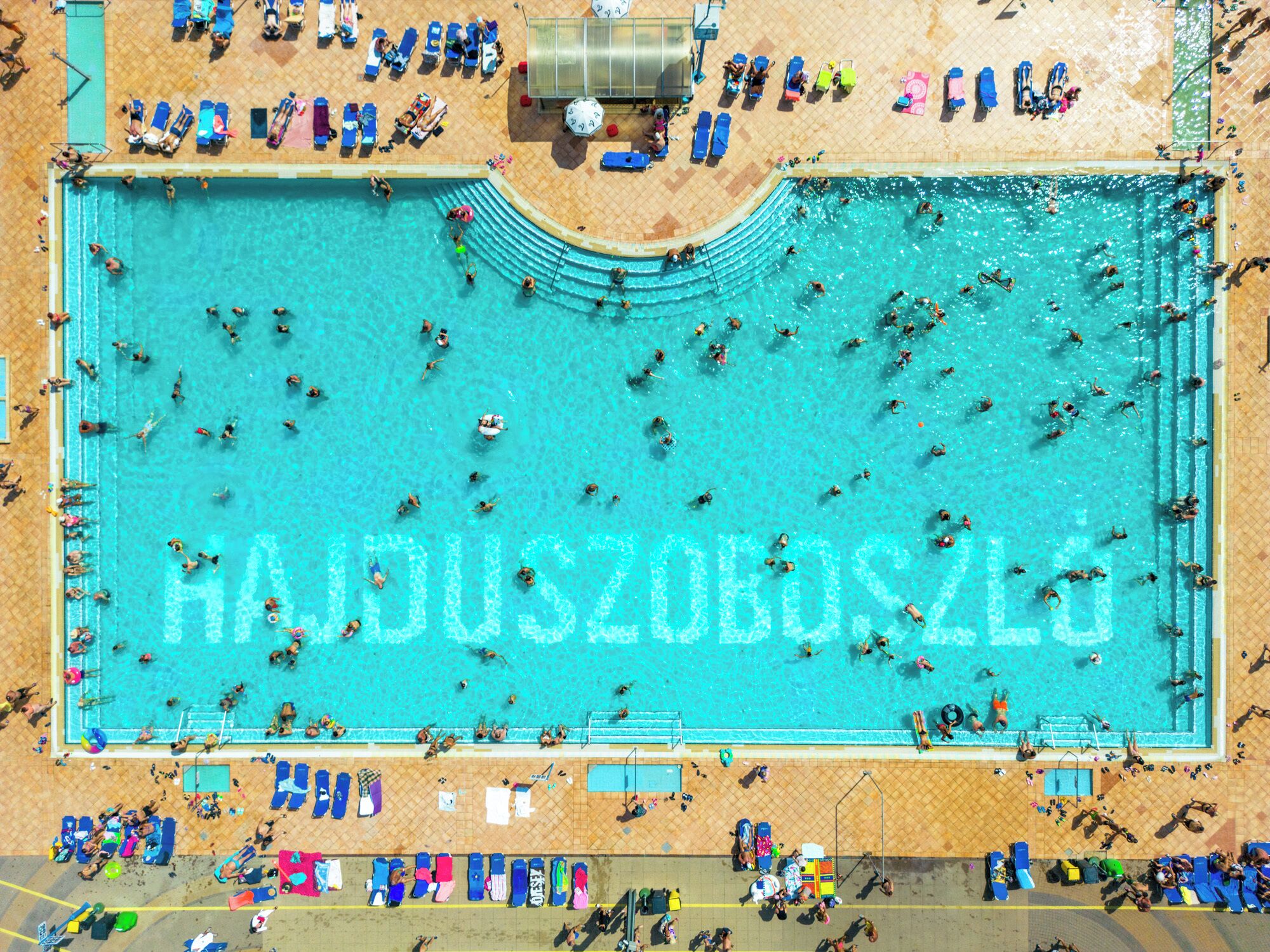 Vista aérea de uma área de piscina com várias pistas, espreguiçadeiras e um espaço de estar coberto.