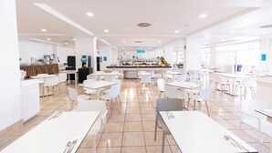 Uma cafeteria ampla com mesas e cadeiras brancas, área de buffet e balcão com diversos tipos de comida.
