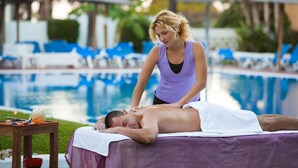 Um homem recebendo massagem de uma mulher em ambiente de piscina, com espreguiçadeiras e guarda-sóis ao fundo.