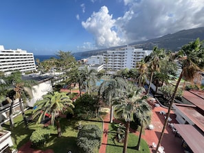 Pátio de hotel com palmeiras, piscina e vista para o oceano e edifícios.