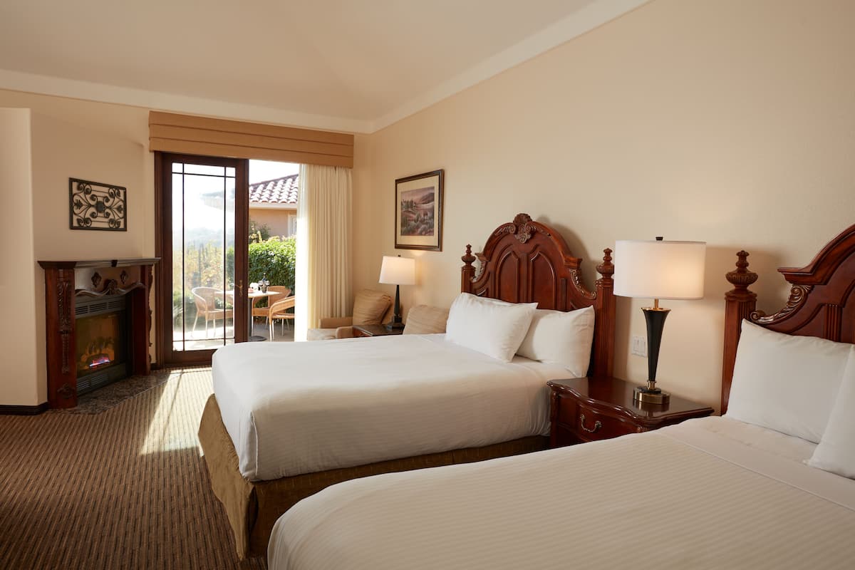 Quarto de hotel com duas camas, lareira, área de jantar com mesa e cadeiras, e vista para o exterior.