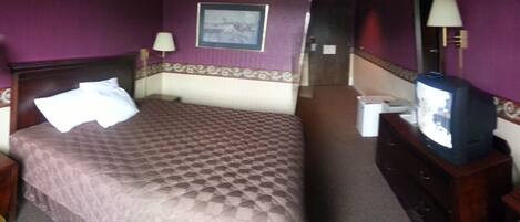 Quarto de hotel com cama, cadeira, escrivaninha e televisão.