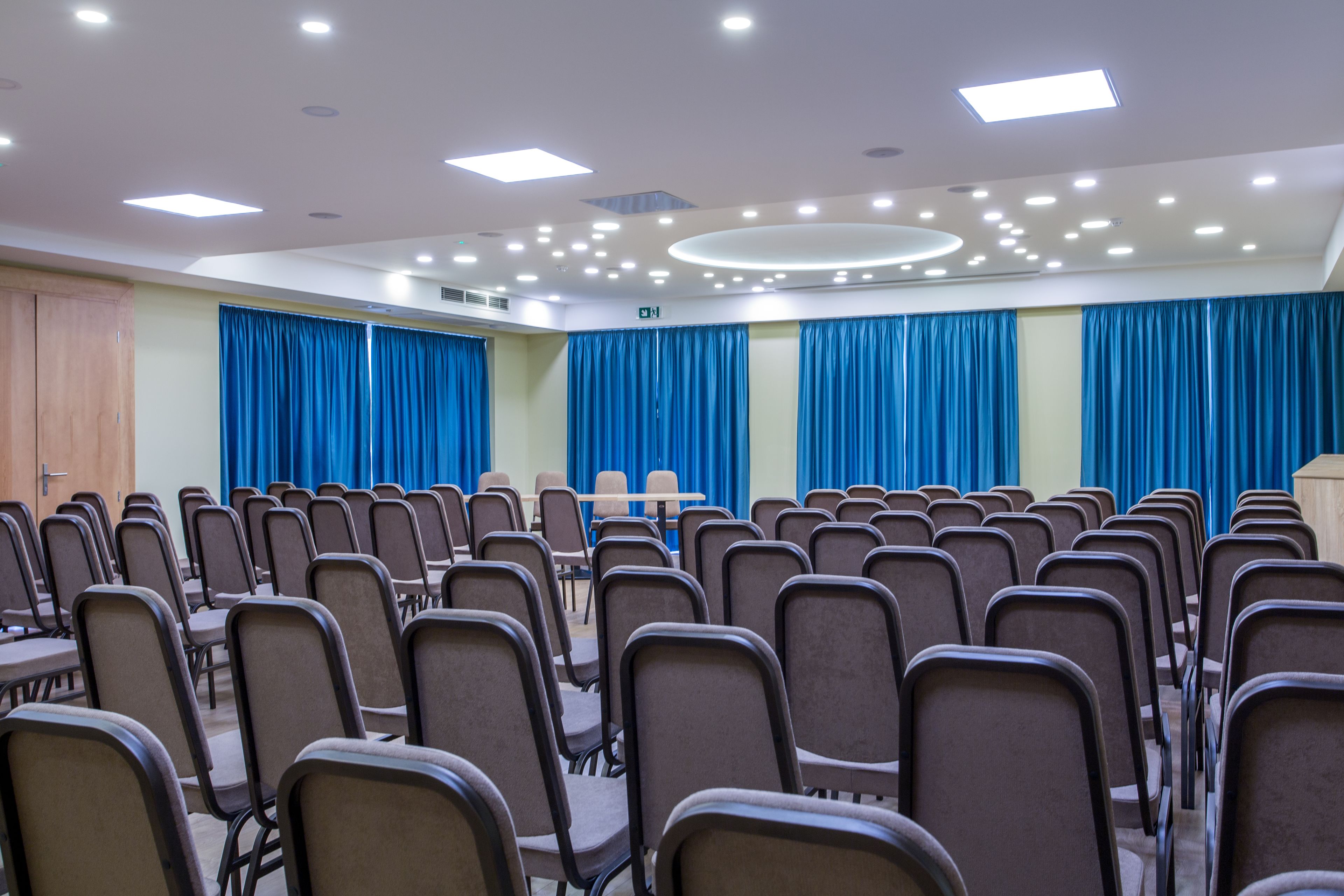Sala de conferência com fileiras de cadeiras, uma mesa e cortinas azuis.