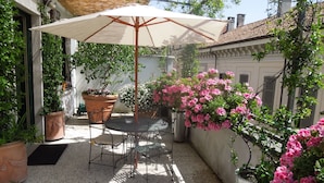 Um pátio com mesa e cadeiras, rodeado por plantas em vasos e um grande guarda-sol.