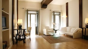 Uma sala de estar espaçosa com um sofá branco, uma mesa de centro de vidro e piso de madeira. Há uma televisão em uma prateleira à esquerda e um abajur em uma mesinha lateral.