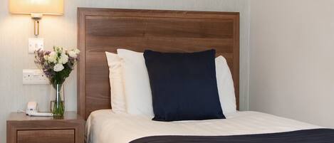 Cama bem arrumada com almofadas brancas e azul-marinho, cabeceira escura e mesa de cabeceira com abajur e um vaso de flores.