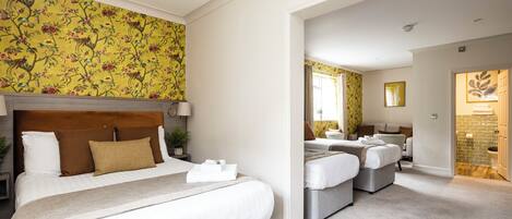 Quarto de hotel com uma cama grande, uma cama menor e um sofá. Inclui uma casa de banho com chuveiro e sanita. O quarto tem papel de parede floral amarelo e uma cabeceira de madeira.