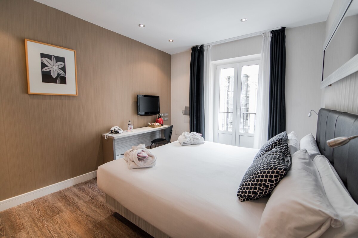 Quarto de hotel com cama, escrivaninha com telefone, televisão e janela com cortinas.