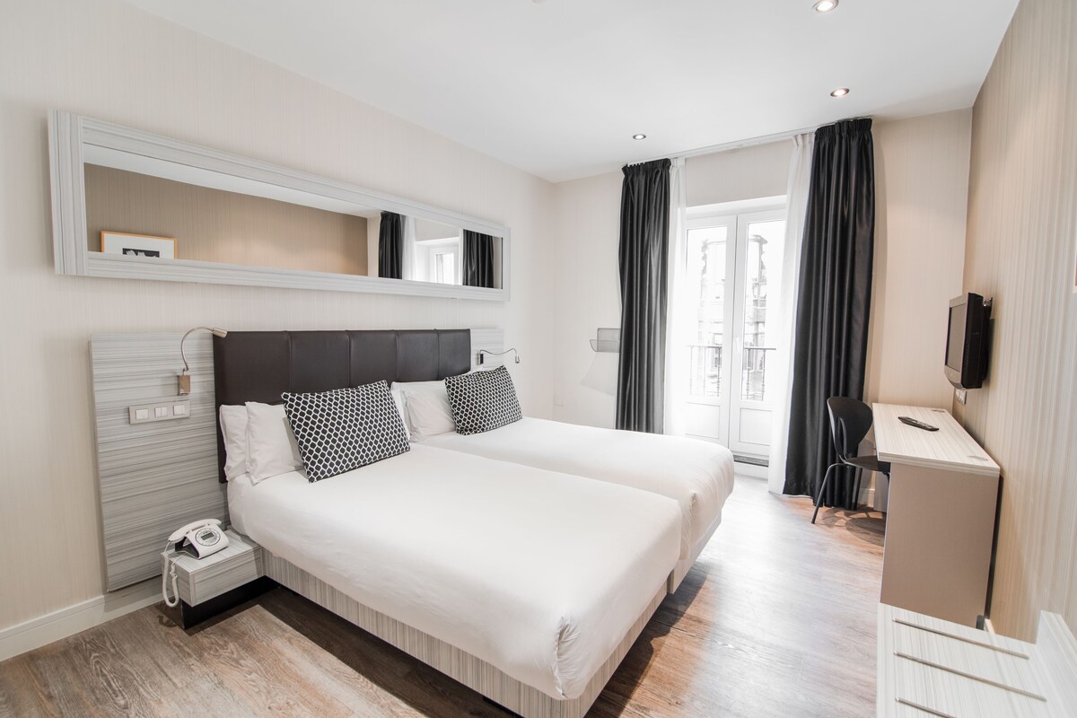 Quarto de hotel com cama de casal, mesas de cabeceira, escrivaninha, cadeira, telefone, espelho e janela com cortinas.