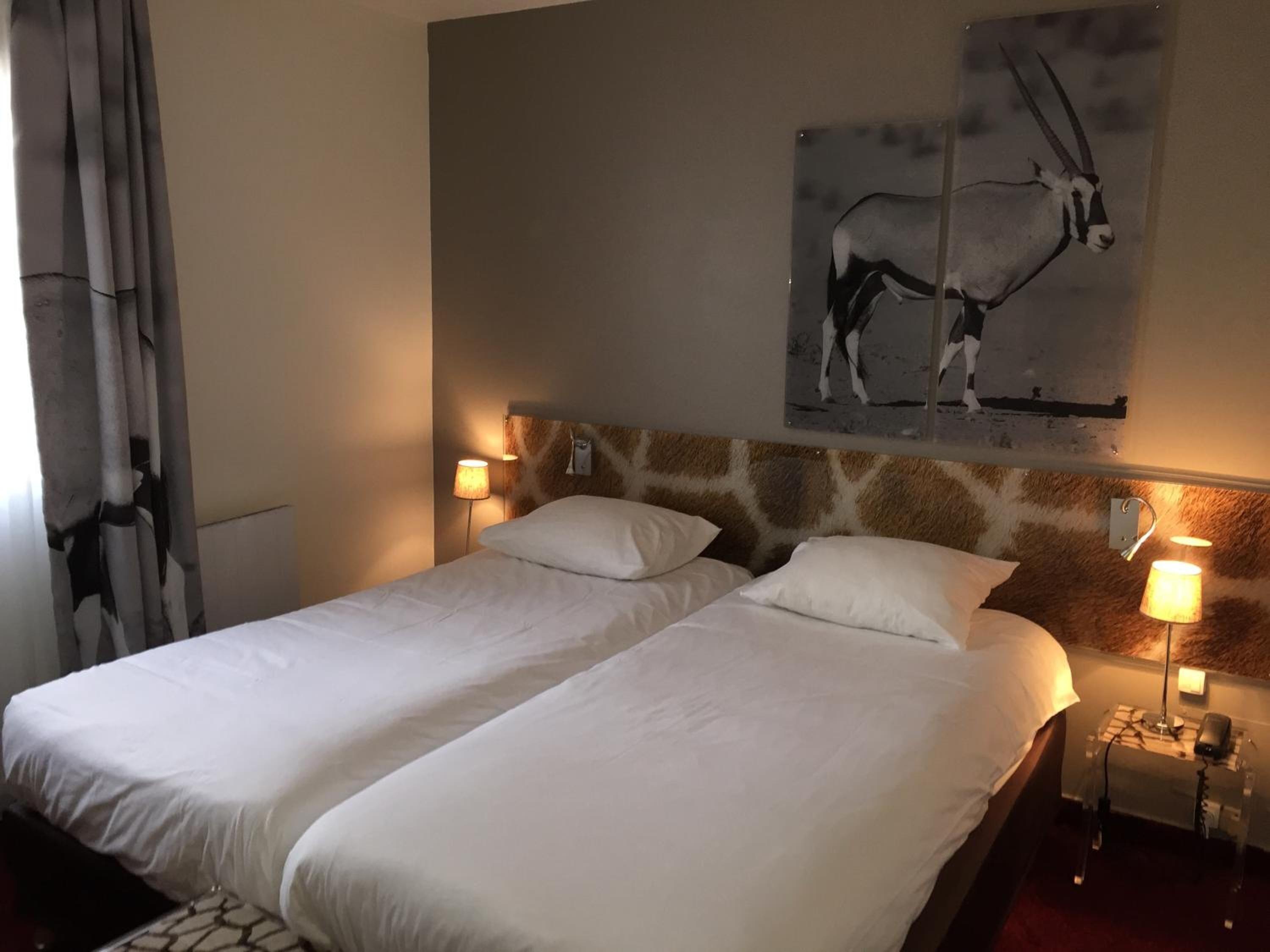 Quarto de hotel com cama de casal, duas luminárias de cabeceira, cabeceira estampada com girafas e uma pintura de girafa na parede.