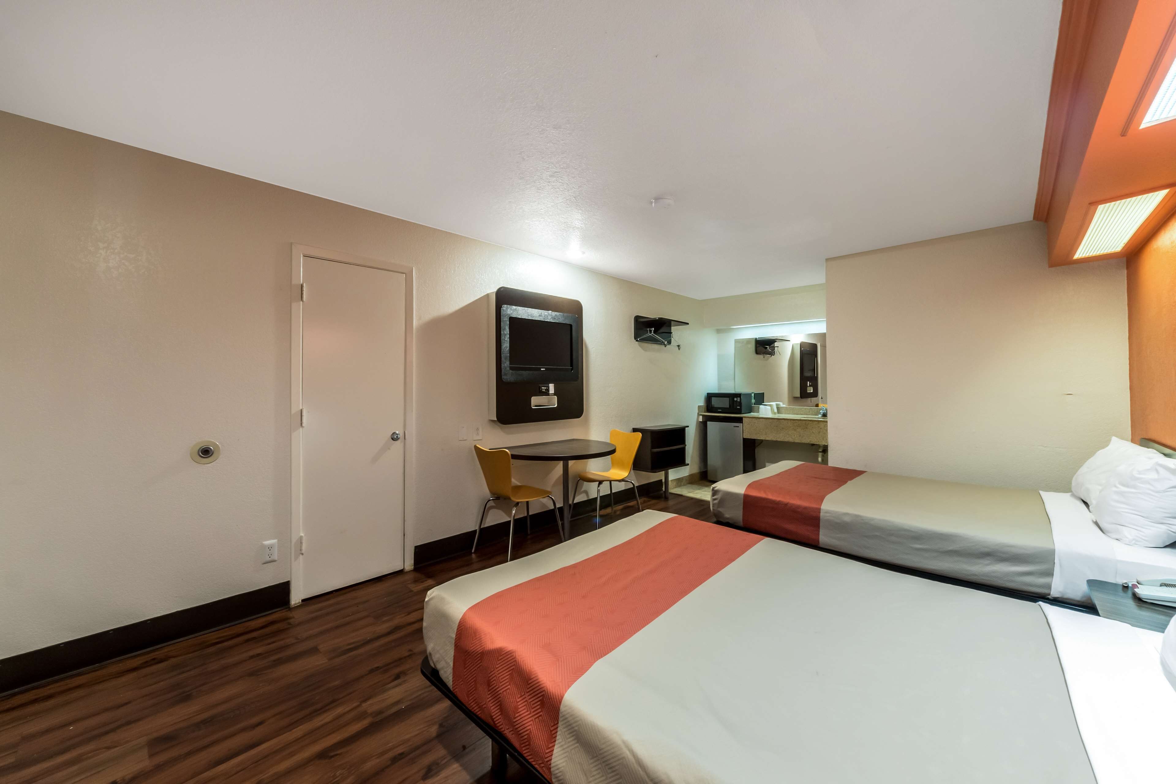 Quarto de hotel com duas camas, uma escrivaninha, uma cadeira, uma televisão e uma pequena cozinha equipada.