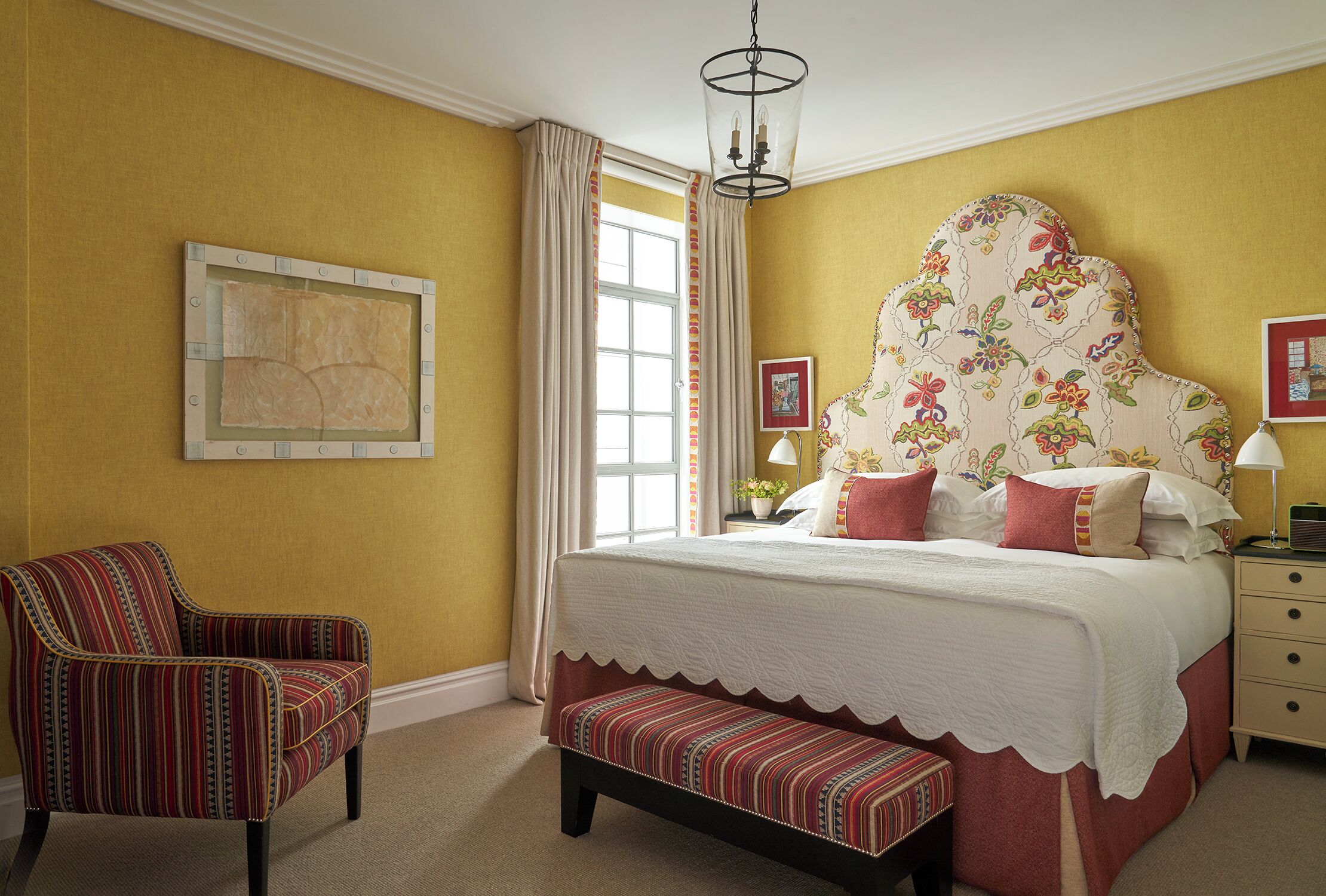 Quarto com cama, cadeira, banqueta, mesa de cabeceira e um quadro na parede.