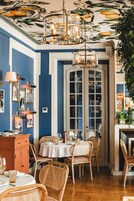 Uma sala de jantar com parede azul, piso de madeira, cadeiras de vime, mesa branca e lustre de vidro.