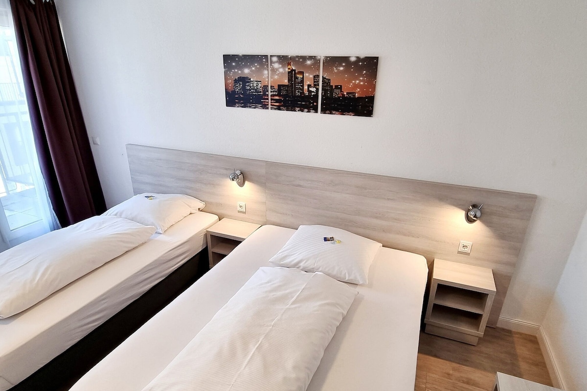 Quarto de hotel com duas camas de solteiro, mesa de cabeceira com luminária, uma obra de arte na parede e janela com cortinas.