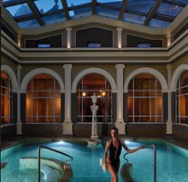 Uma grande piscina interior com teto de vidro, janelas em arco e uma fonte central.