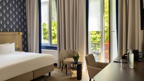 Quarto de hotel com uma cama, uma cadeira, uma mesinha, um vaso de planta e vista para o exterior.