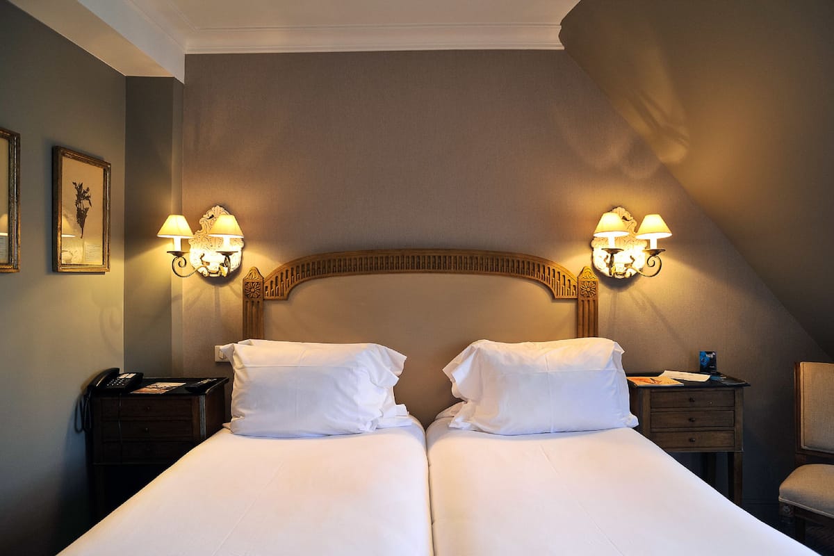 Uma cama de casal com lençóis brancos, duas luminárias de cabeceira, um painel de madeira na cabeceira e uma mesinha de cabeceira com um telefone, além de um quadro na parede.