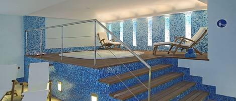 Uma área de piscina interna moderna, com plataforma flutuante, escadas de madeira e cadeiras de descanso.